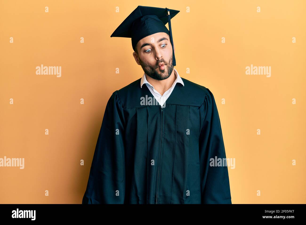 Young hispanic man wearing graduation cap and ceremony robe making fish ...