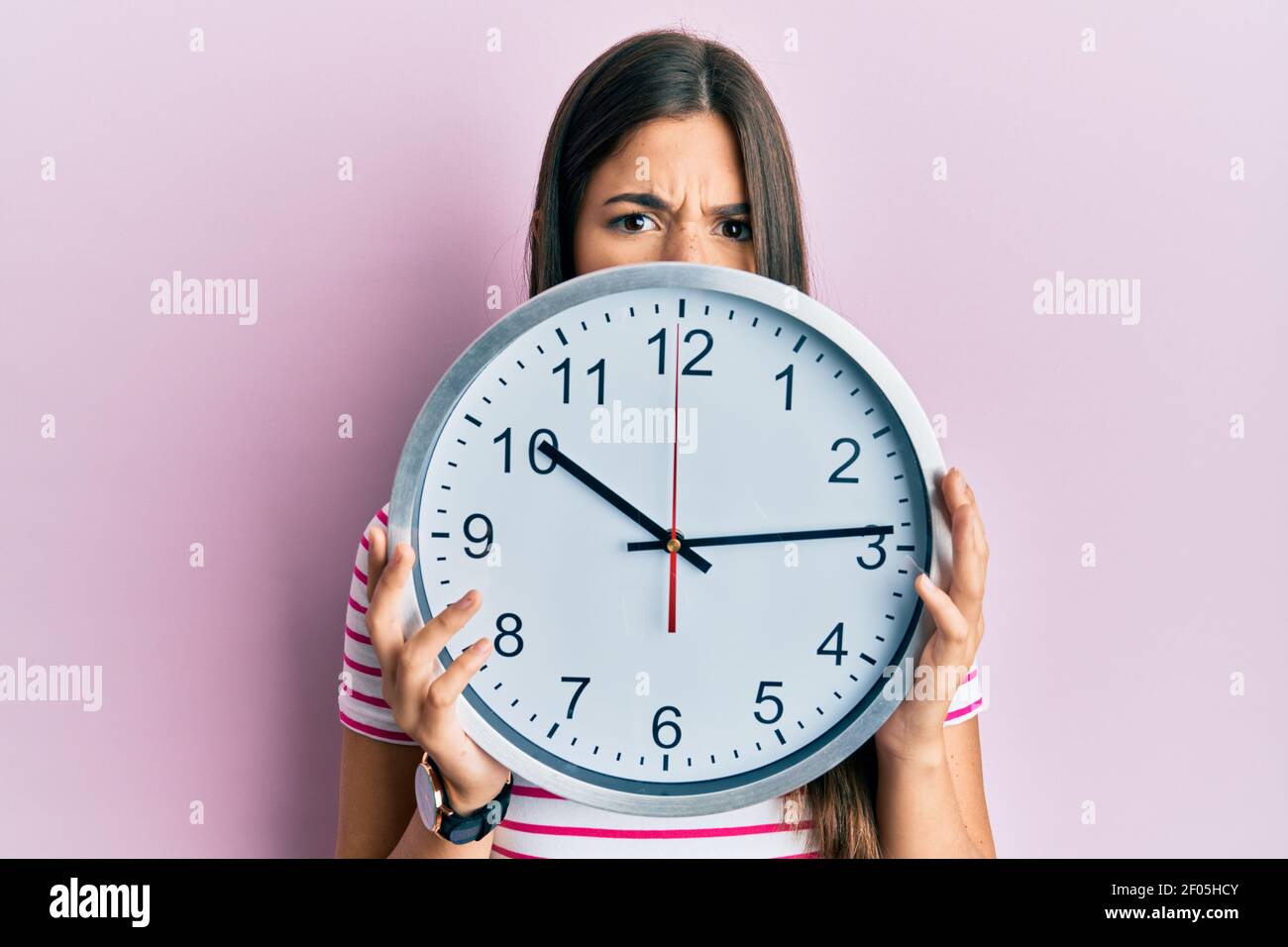 Young brunette woman holding big clock covering face skeptic and ...