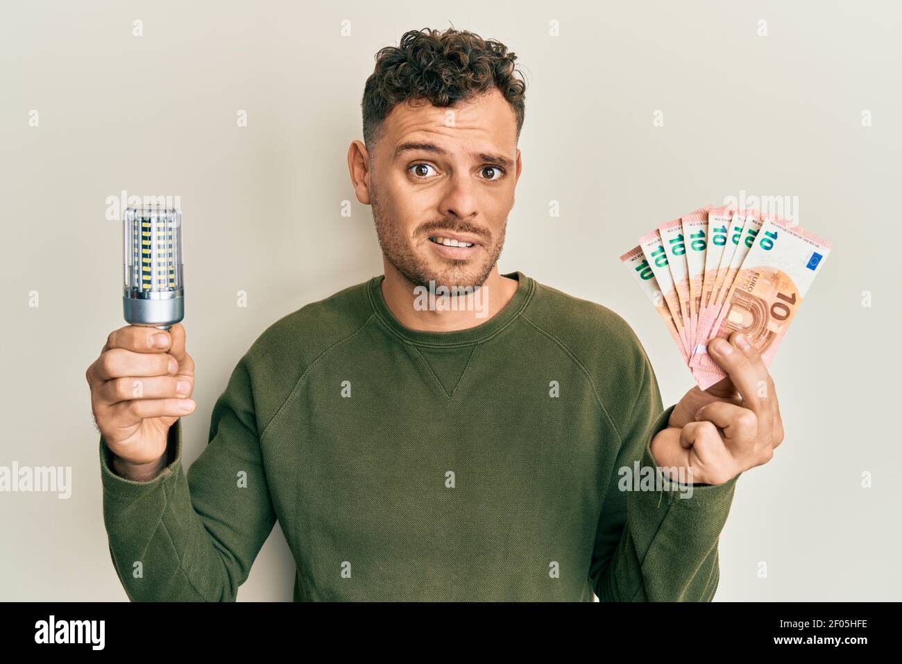 Young hispanic man holding led bulb and euros banknotes clueless and ...