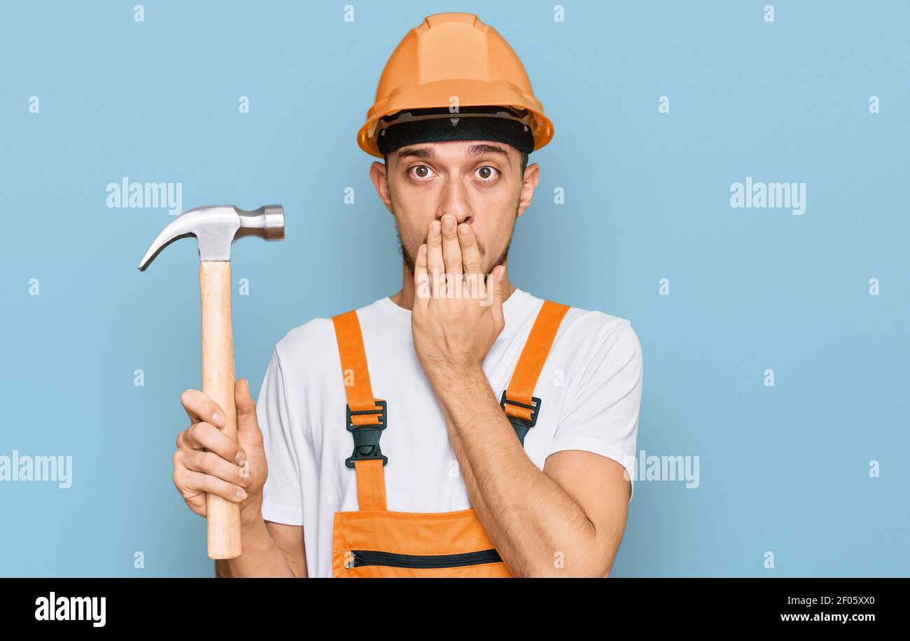 Hispanic young man wearing hardhat holding hammer covering mouth with ...