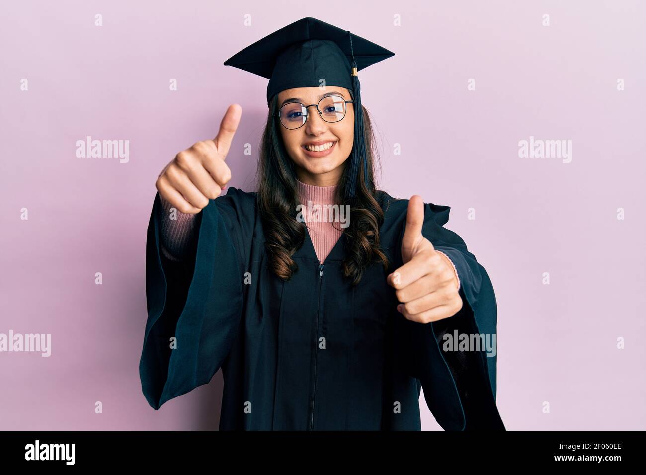 Young hispanic woman wearing graduation cap and ceremony robe approving ...
