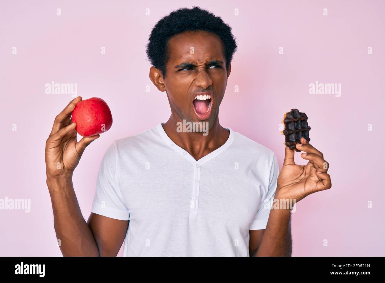 Handsome african man holding red apple and chocolate waffle angry and ...