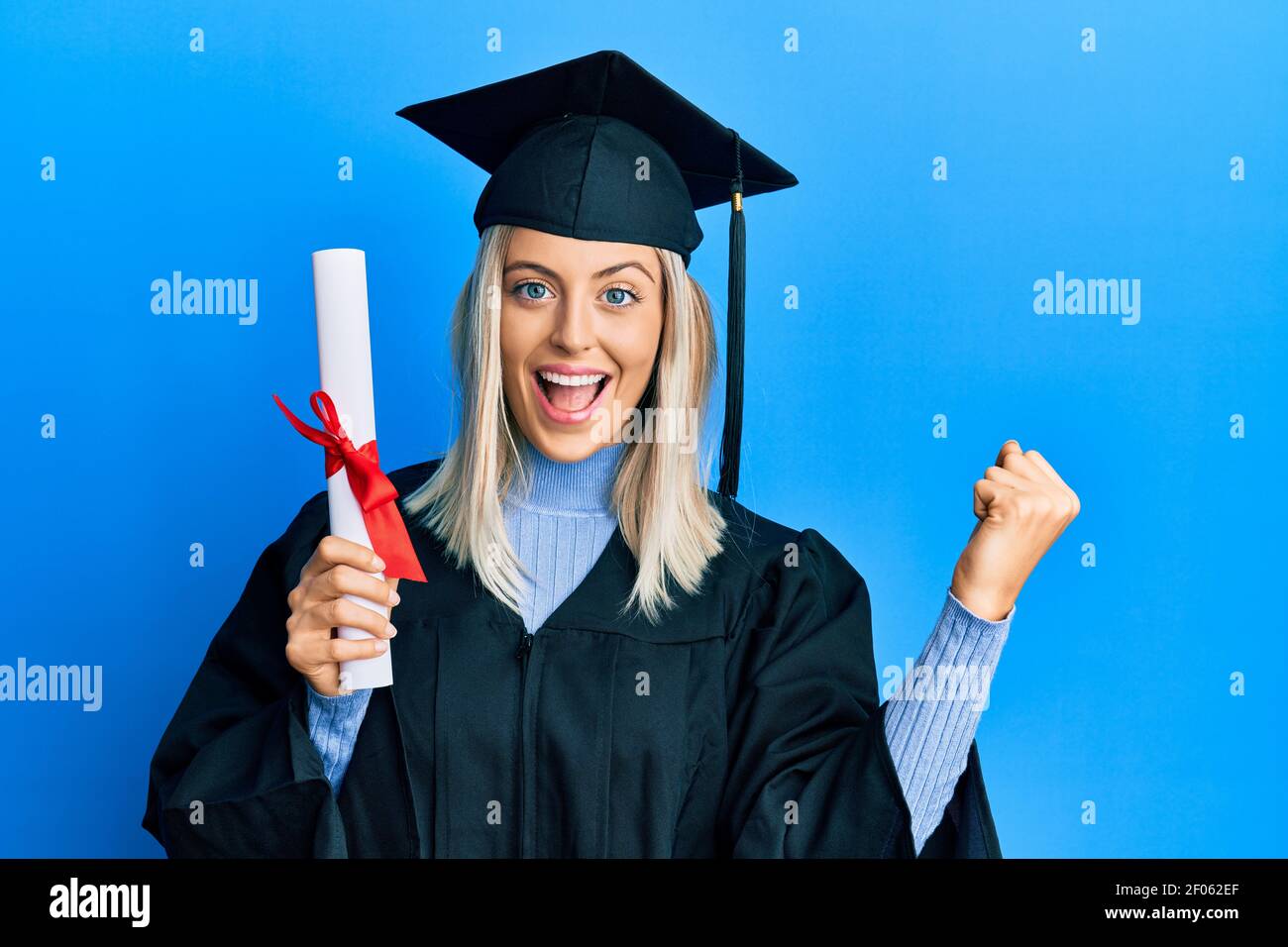 Beautiful blonde woman wearing graduation cap and ceremony robe holding ...