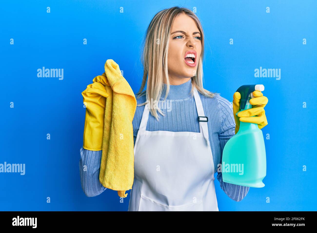 Beautiful blonde woman wearing cleaner apron holding cleaning products ...