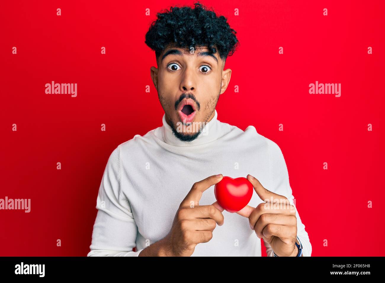 Young arab handsome man holding heart afraid and shocked with surprise ...
