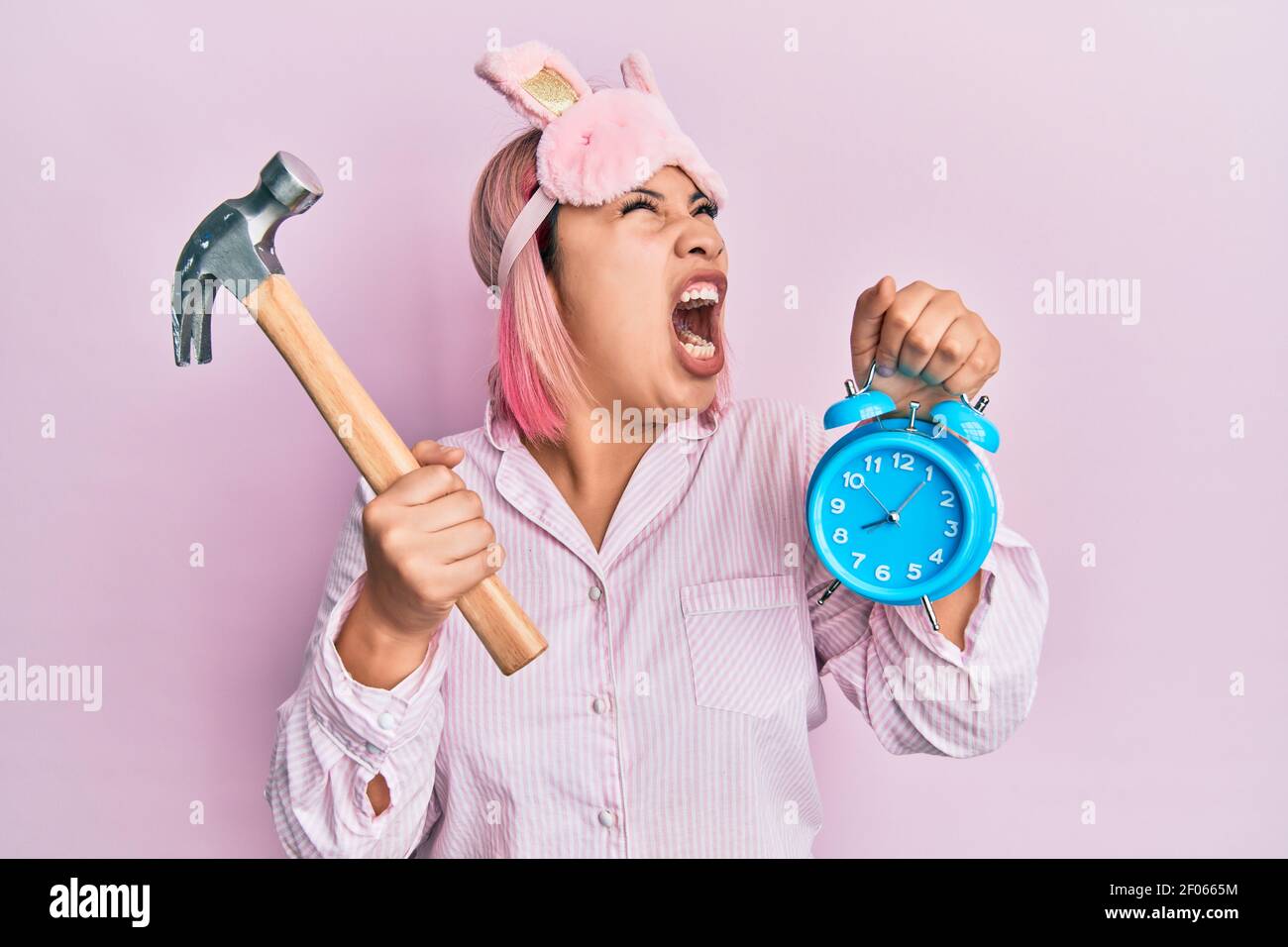 Hispanic woman with pink hair wearing pajama holding hammer and alarm ...