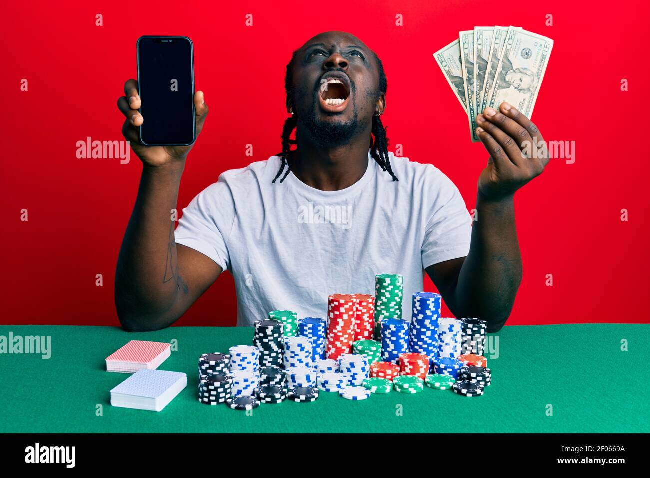 Handsome young black man playing poker holding smartphone and dollars ...