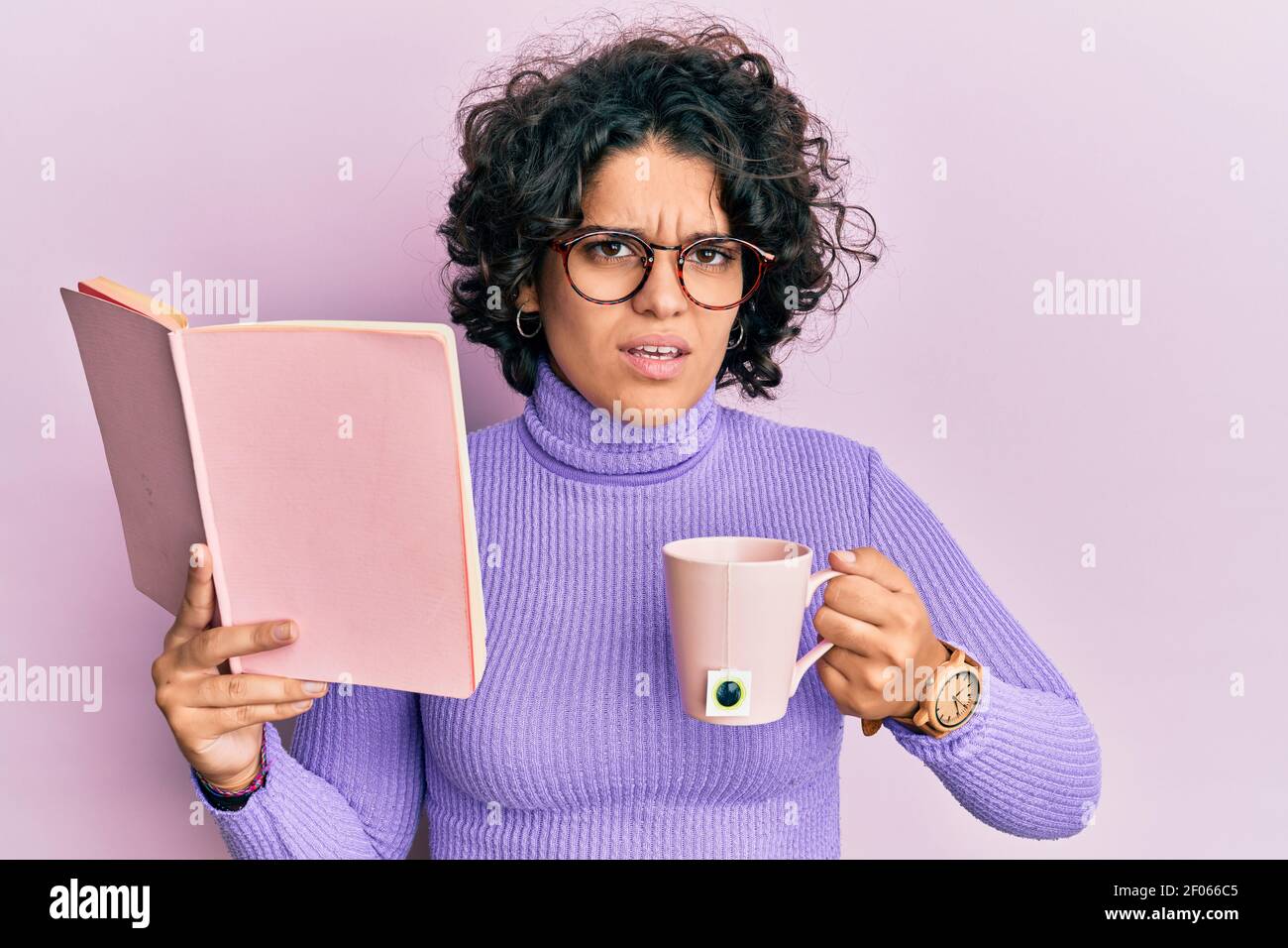 Young hispanic woman with curly hair reading a book and drinking a cup ...