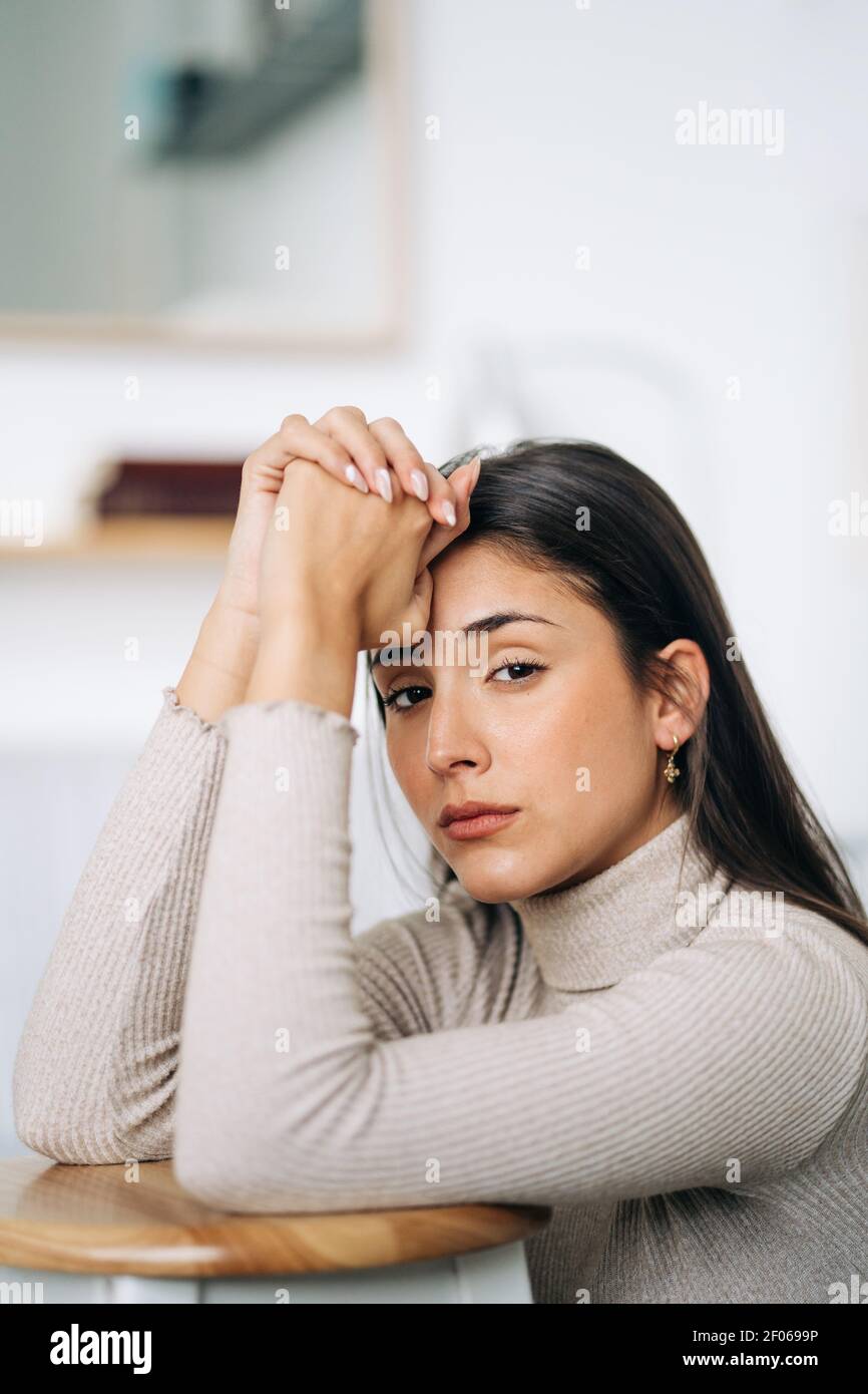 Unemotional young thoughtful female looking at camera leaning on stool ...