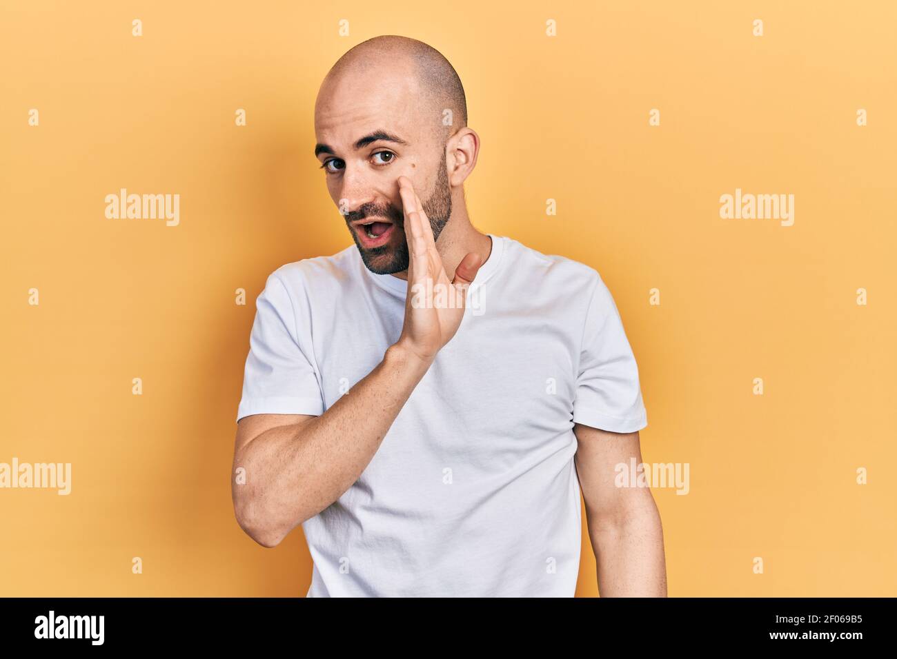 Young bald man wearing casual white t shirt hand on mouth telling ...
