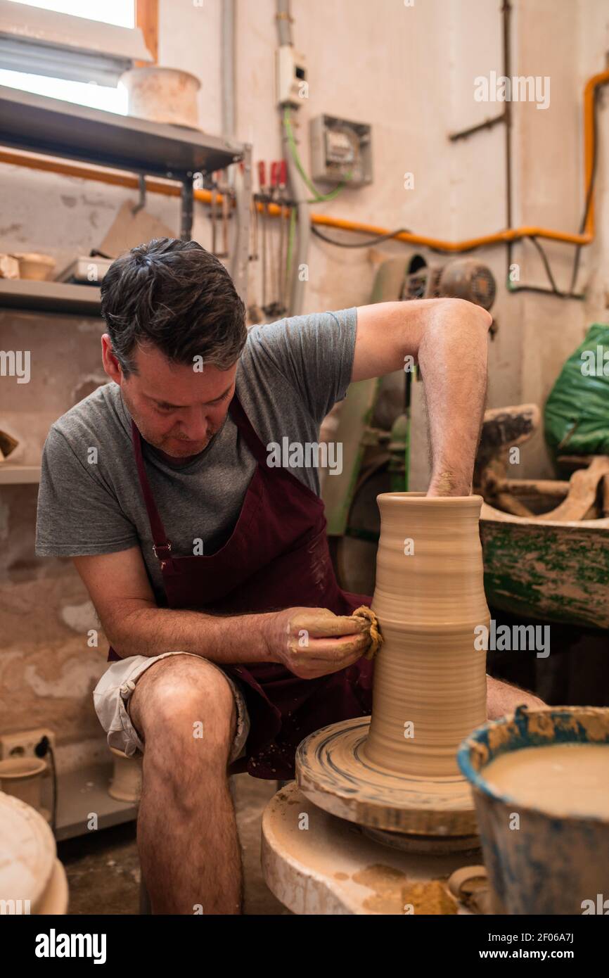 Full body of concentrated male master in apron sitting at table while ...