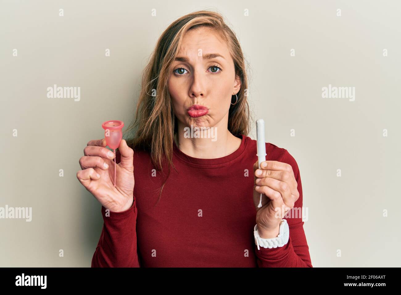 Young blonde woman holding menstrual cup and tampon depressed and worry ...