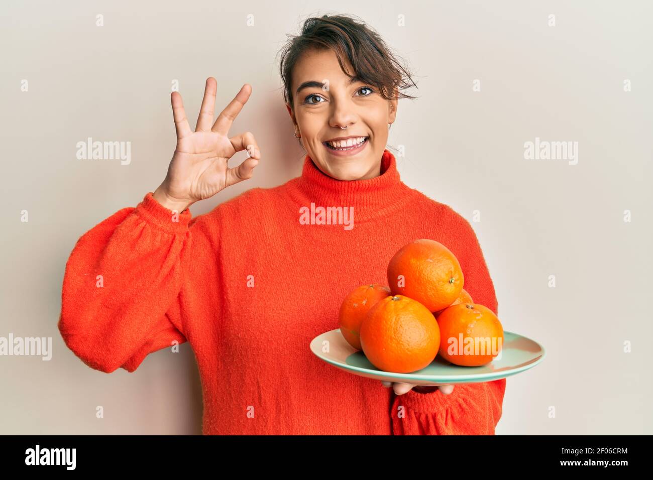 Young hispanic woman holding fresh oranges doing ok sign with fingers ...