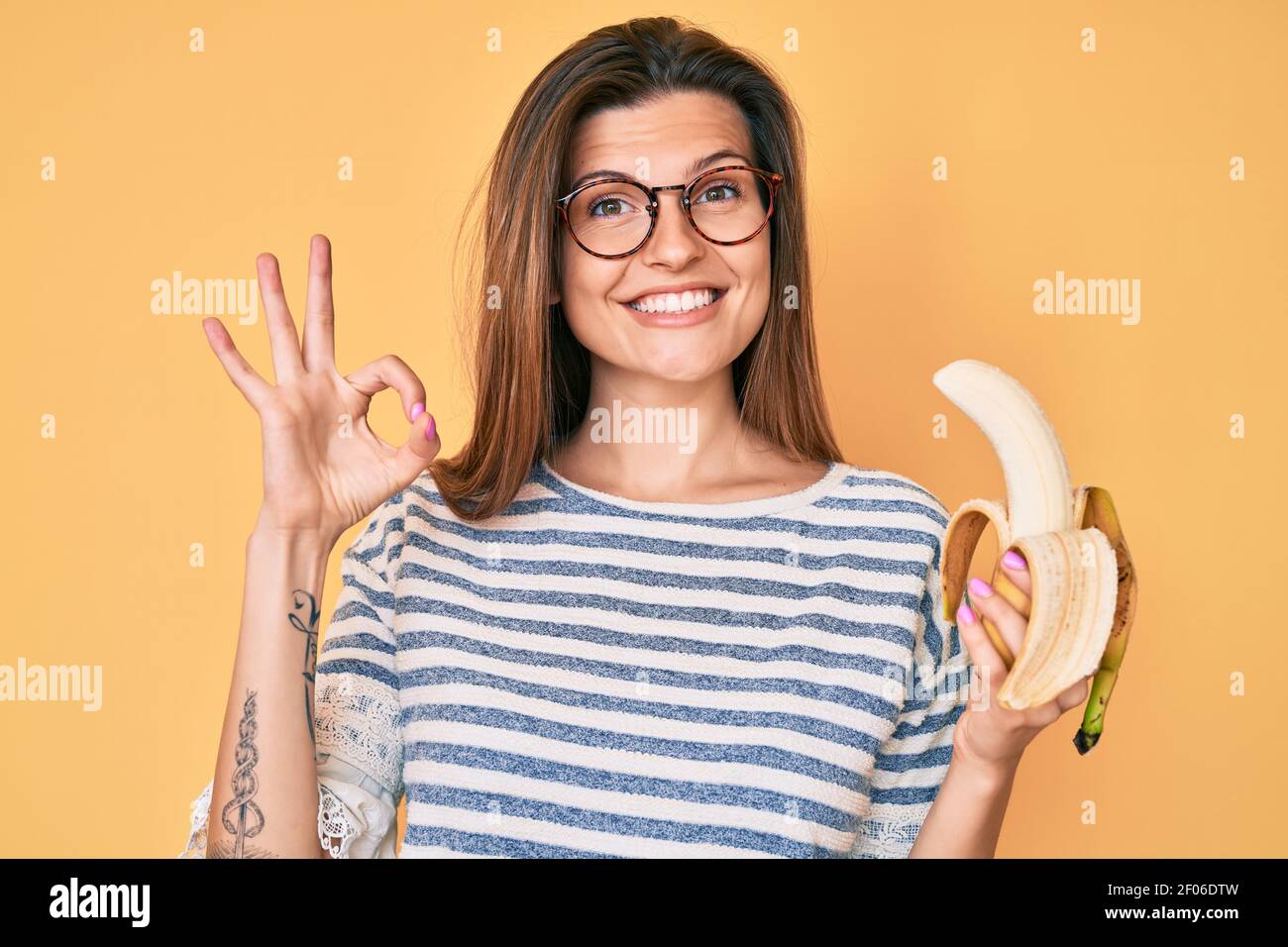 Beautiful caucasian woman eating banana as healthy snack doing ok sign ...