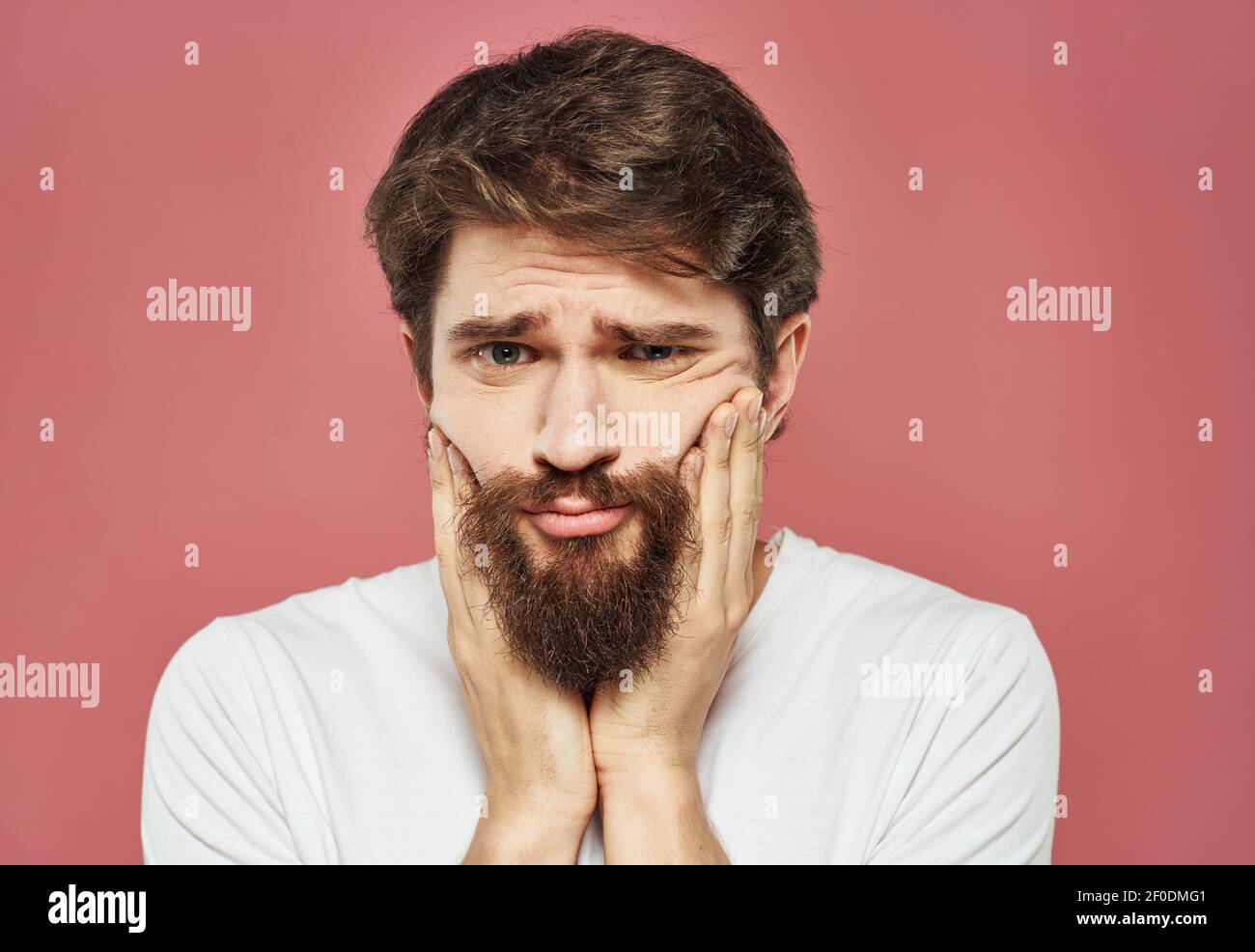 A man with a beard touches his face with his hand on a pink background ...