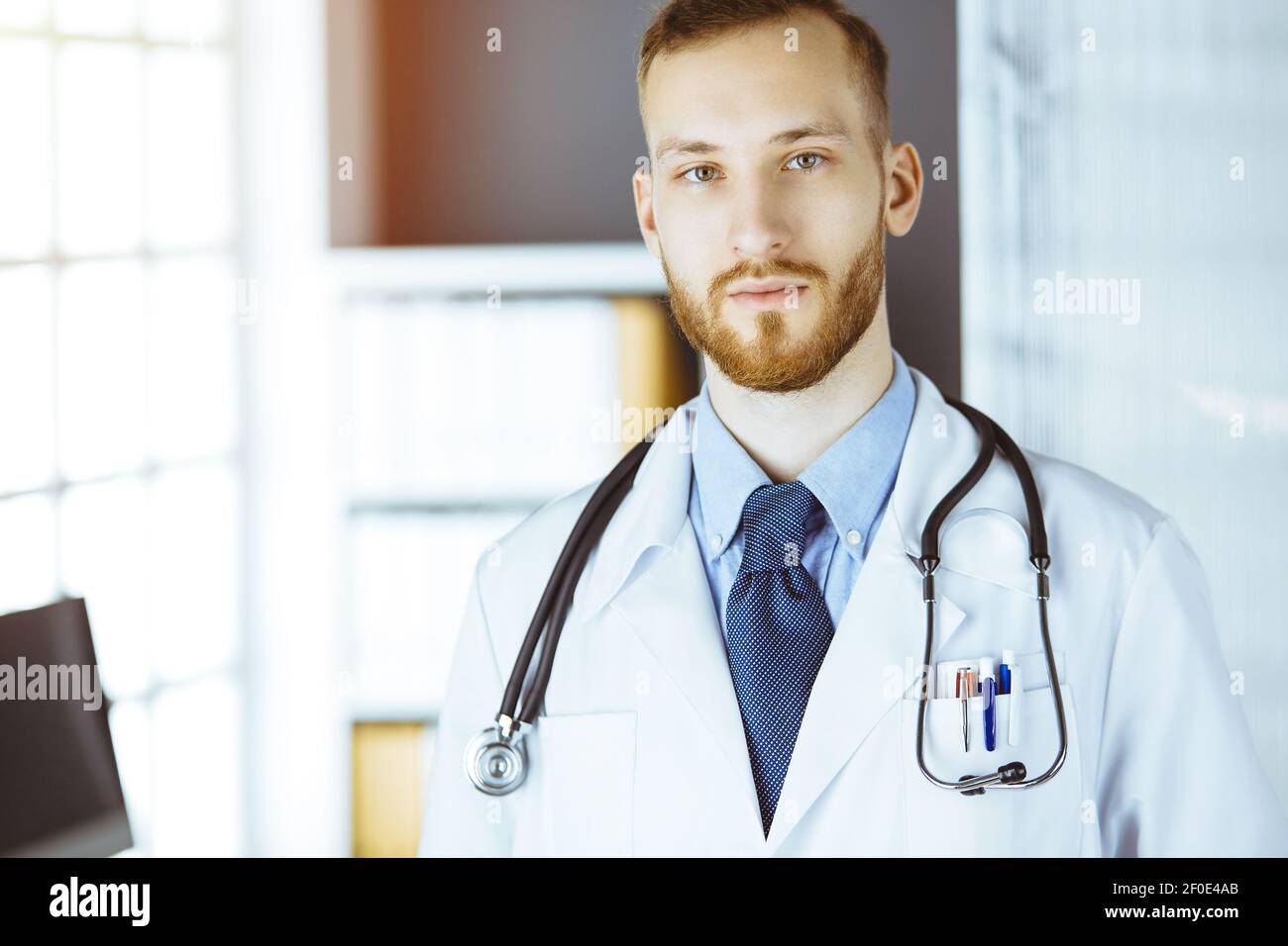 Red-bearded doctor standing straight in sunny clinic near his working ...