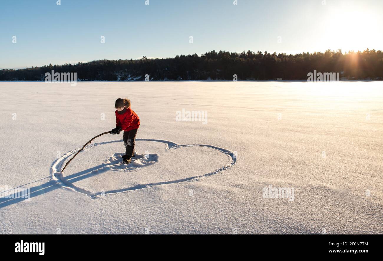 Child drawing a heart shape in an open snowy field in the morning sun ...