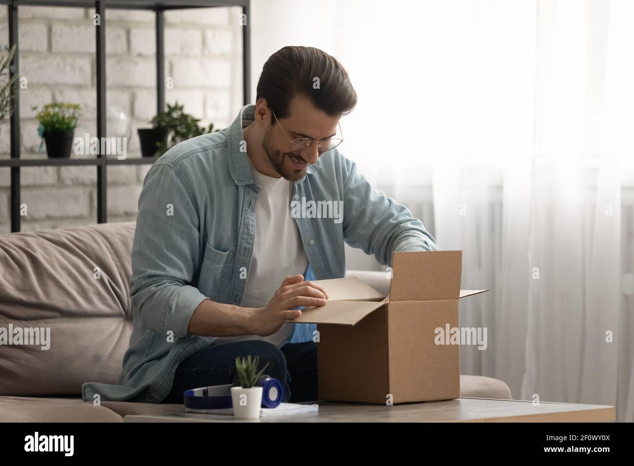 Smiling young guy opening package with consumer goods with pleasure ...