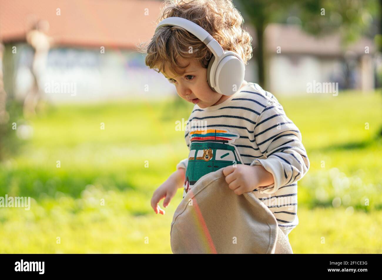 two-year-old boy with headphones playing with a hat in a park in spring ...