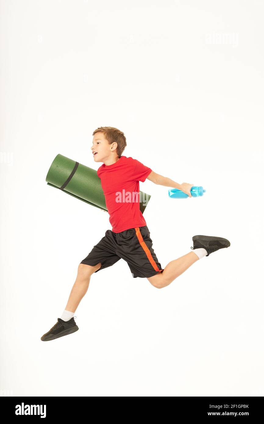 Smiling male kid holding rolled exercise mat and refreshing sports ...