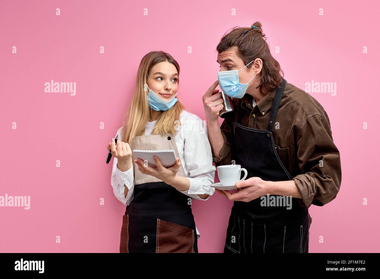 two young waiters in apron and mask discuss orders, ready to serve ...