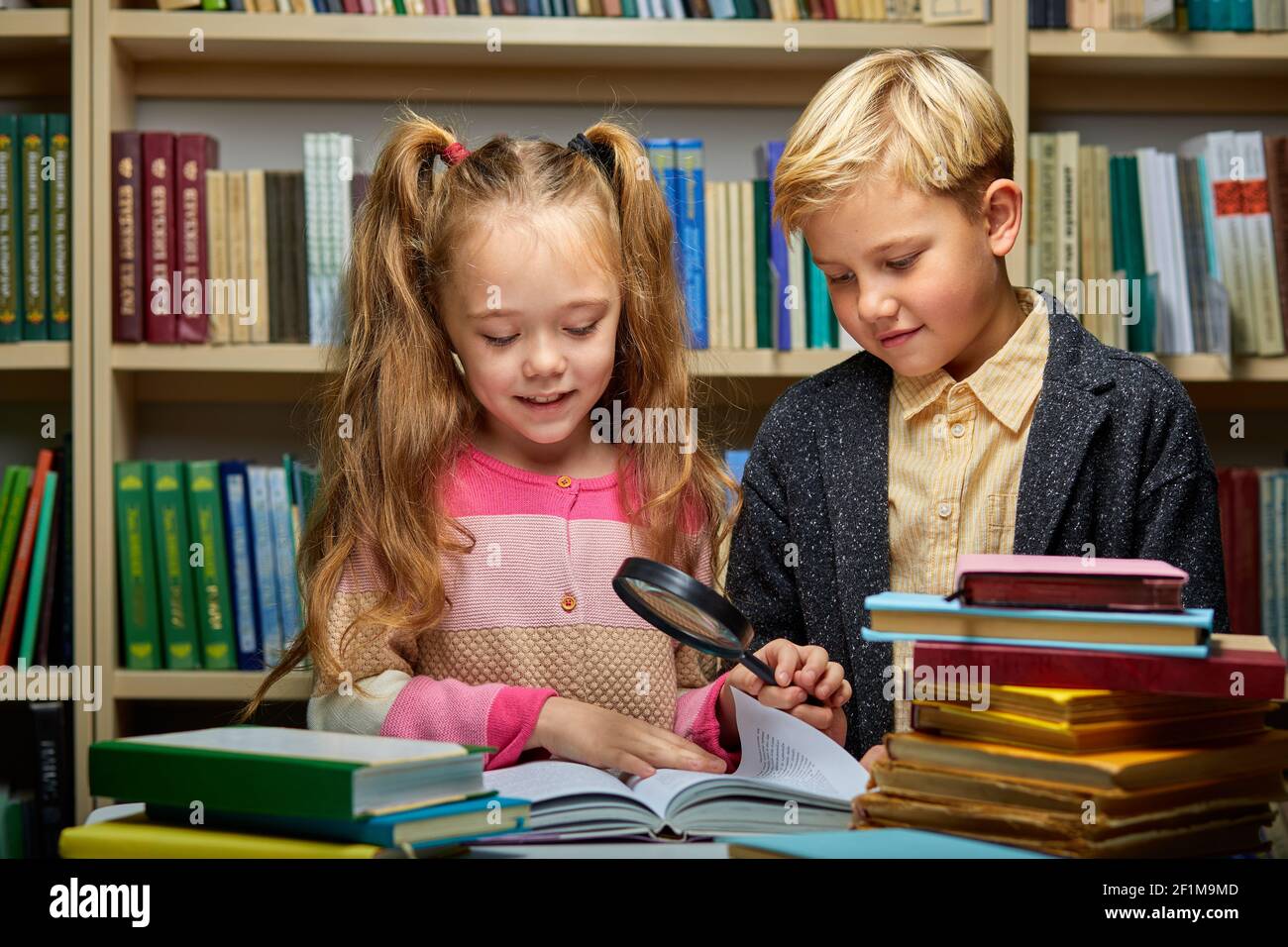 diligent pupil with books in library, boy and girl use magnifying glass ...