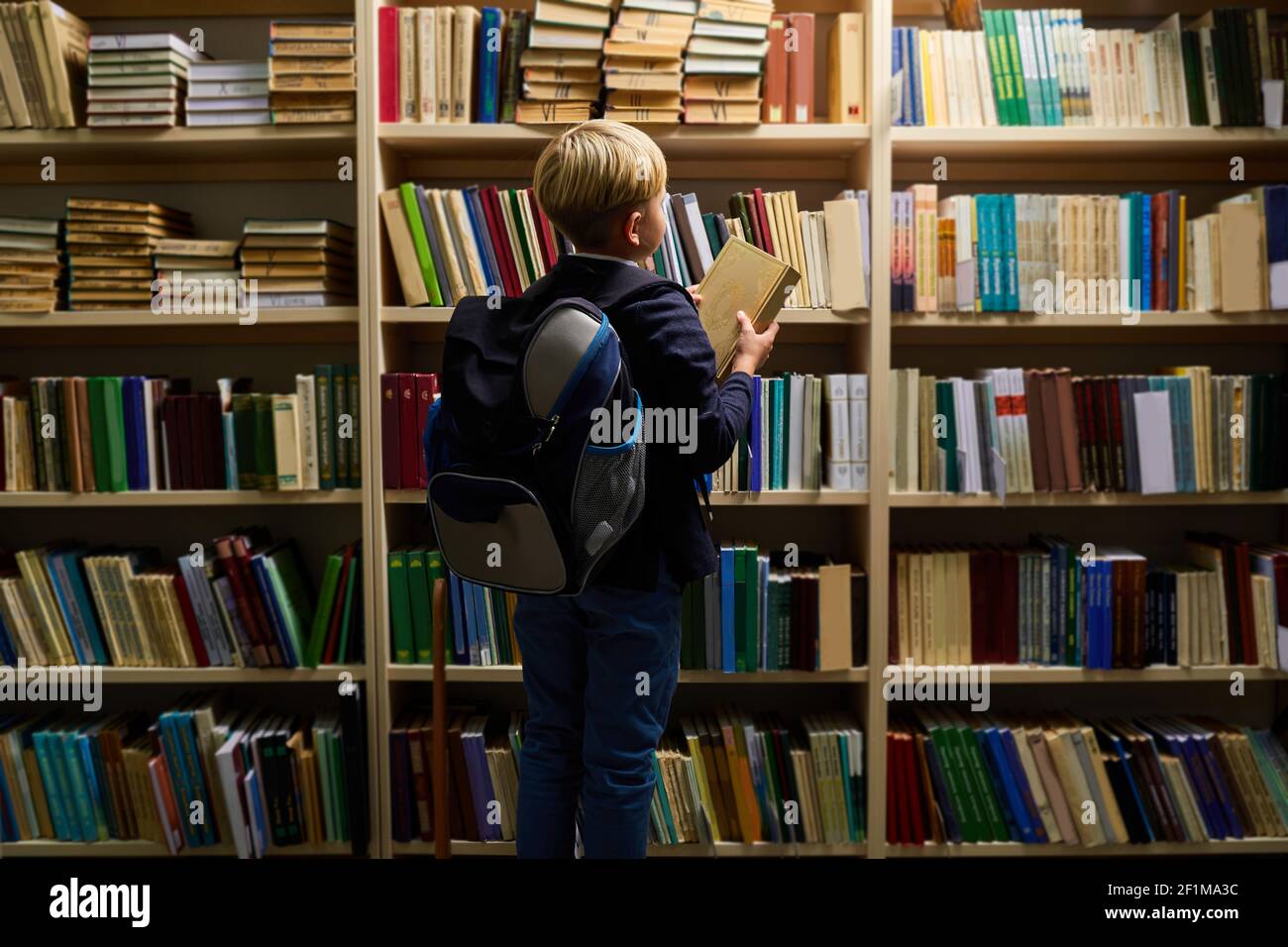 rear view on school boy taking a book from shelves, choosing the book ...