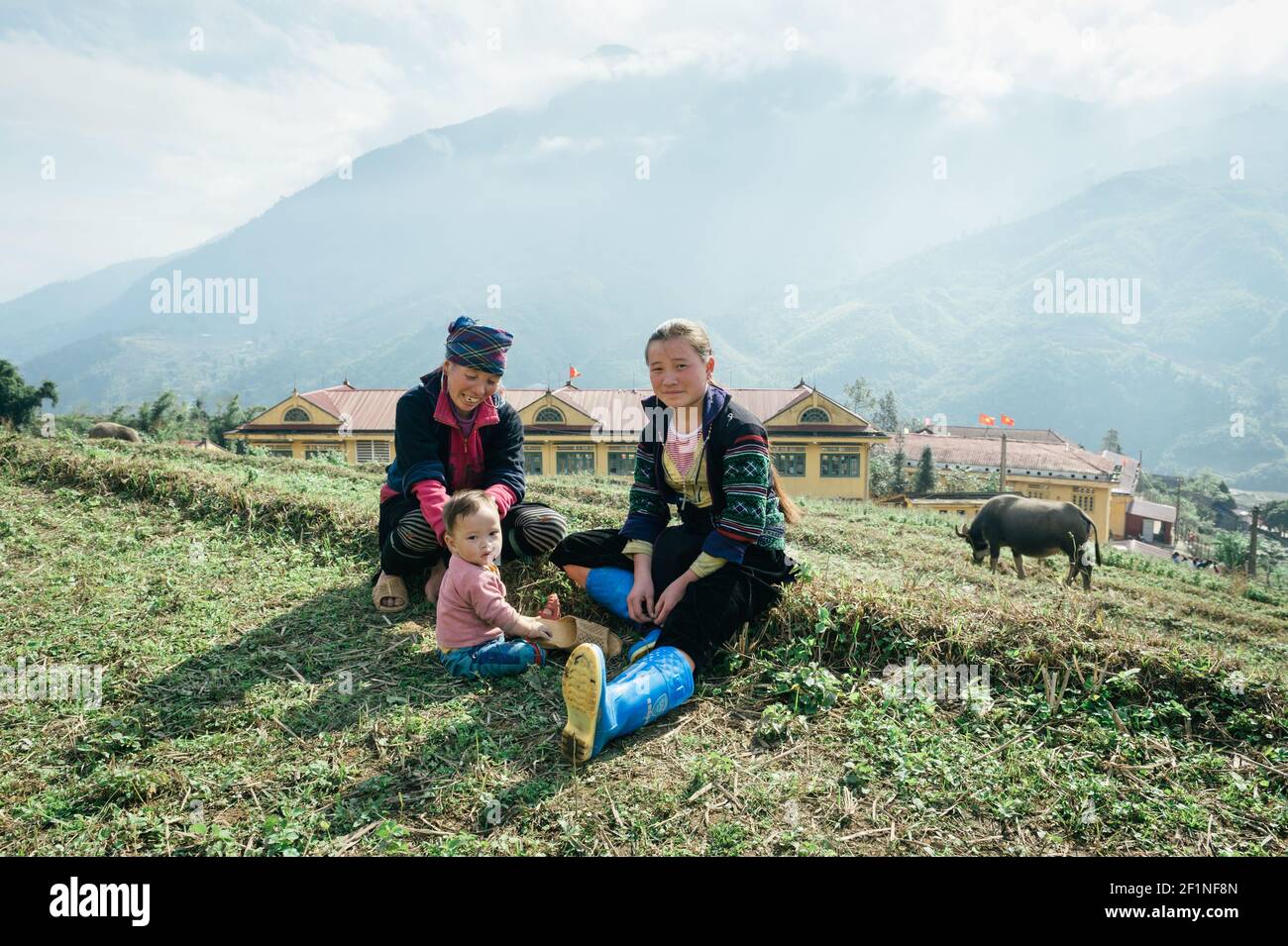 Two Hmong tribe women and a baby, are sitting on the ground in a green ...