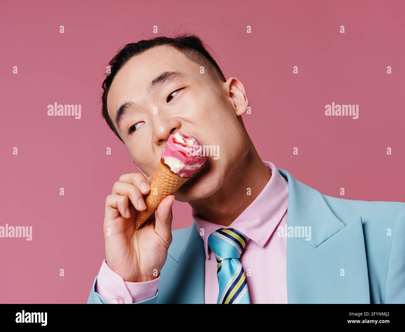 Asian man in a suit eating ice cream in a cone on a pink background ...
