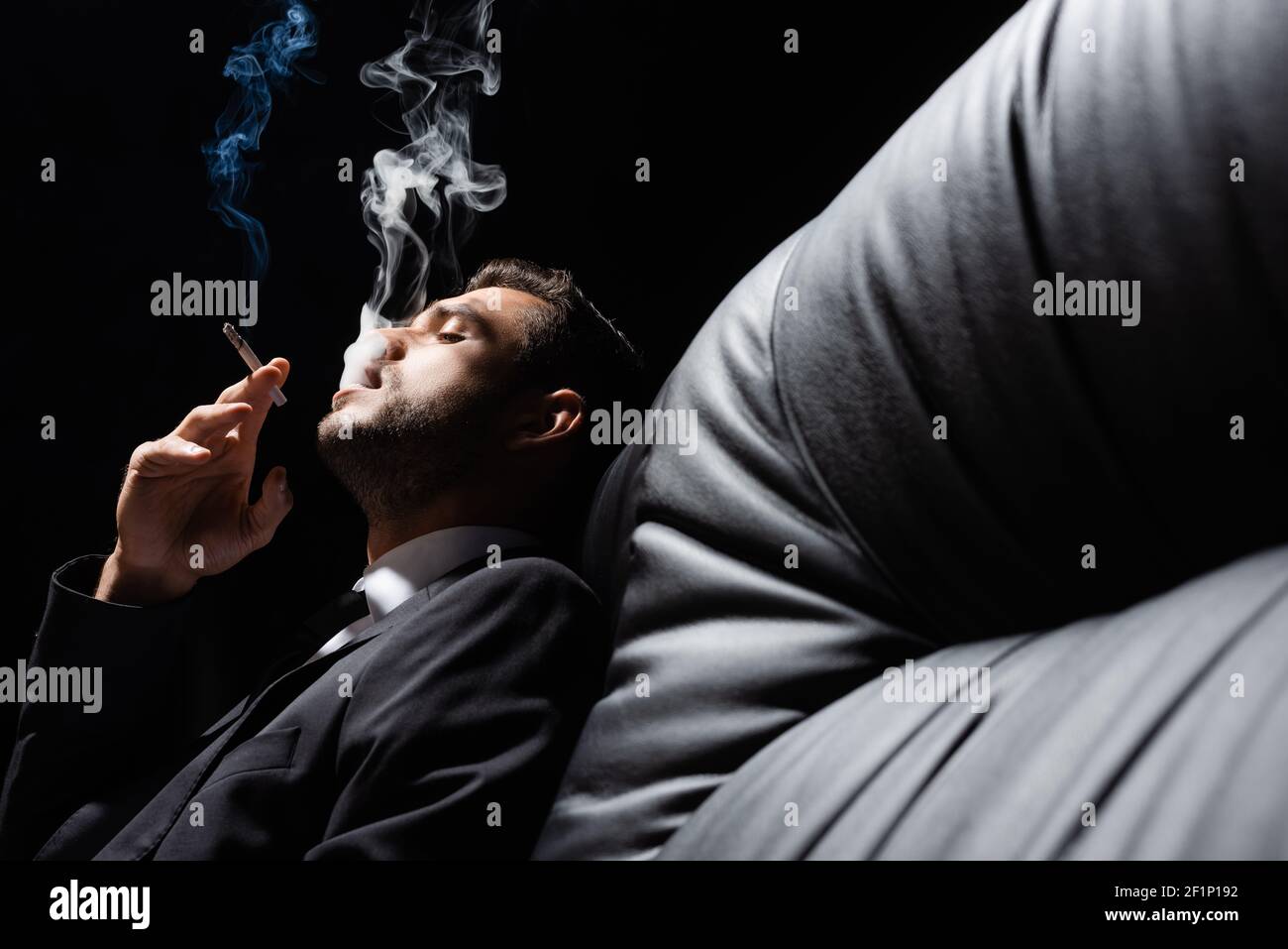 Man in formal wear smoking cigarette on black couch isolated on black ...