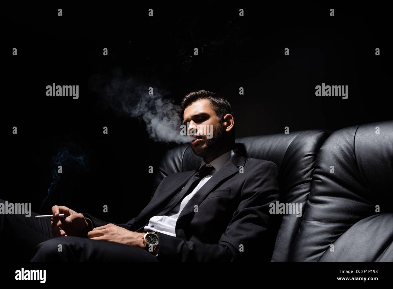Brunette man in formal wear smoking on leather couch isolated on black ...