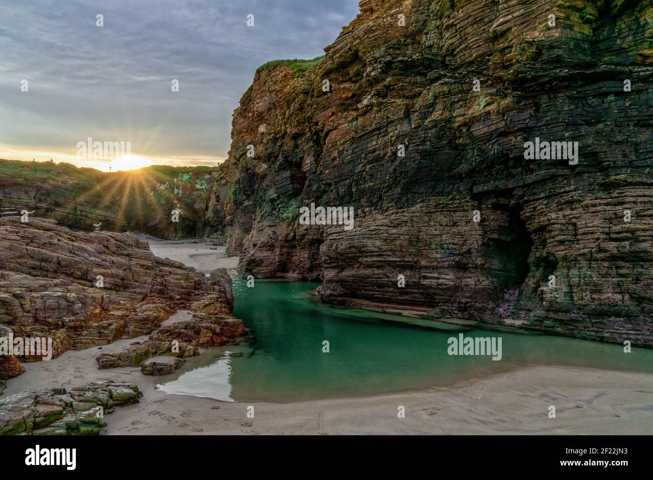 The sun peaks over the edge of a cliff with sandy beach and tidal pools ...
