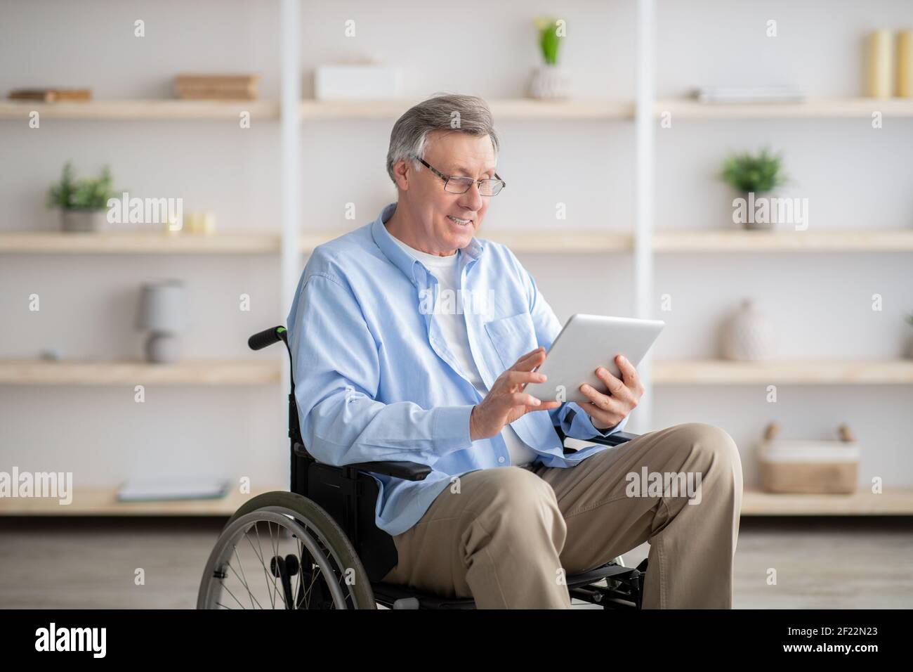 Joyful impaired senior man in wheelchair using tablet computer at home ...