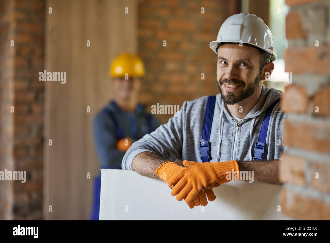 Portrait of positive, handsome young male builder in hard hat smiling at camera, holding drywall ...