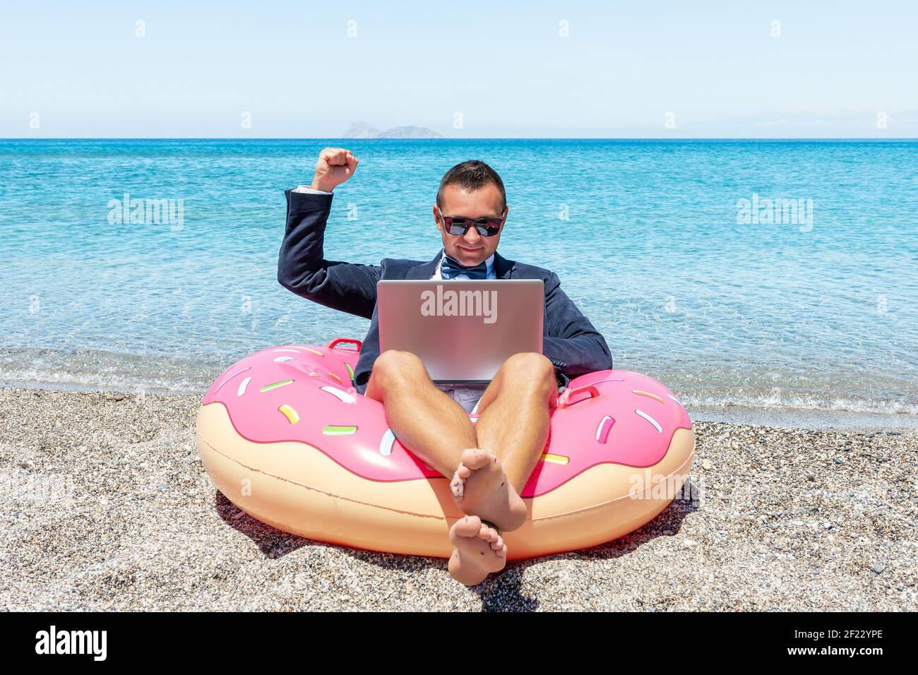 Happy businessman with laptop on inflatable donut on tropical beach ...
