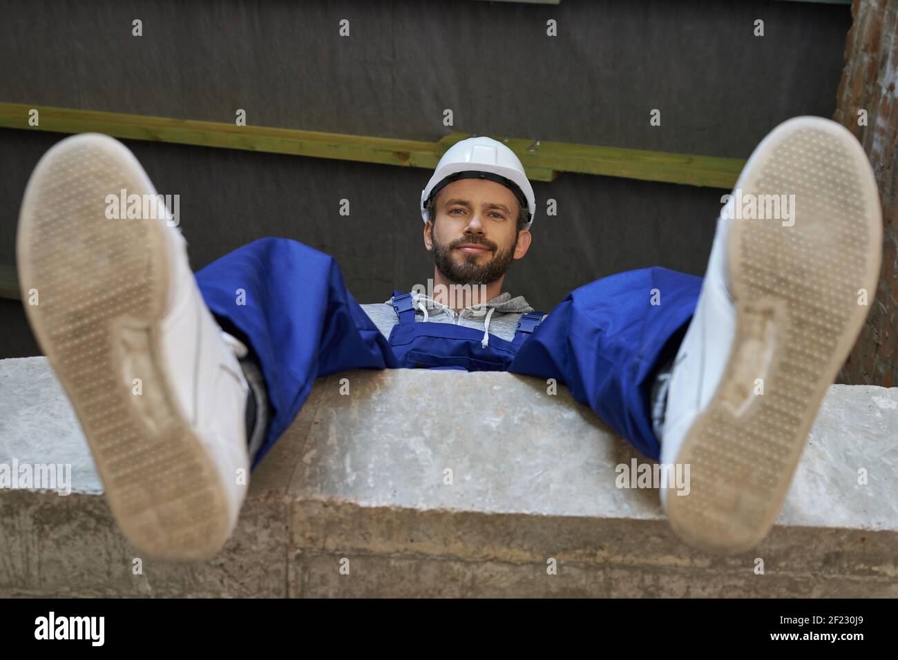 Happy handsome young male builder in blue overalls and hard hat looking ...