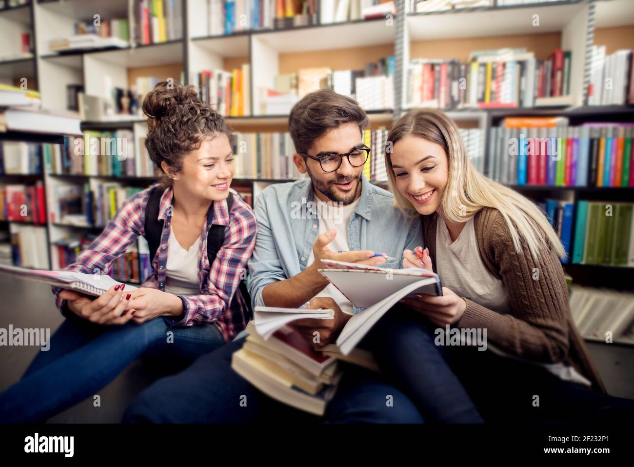 Three young joyful college friends sitting on the library couch getting ...
