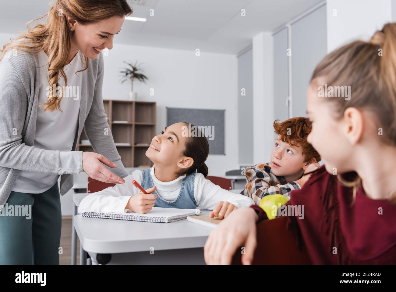 happy teacher and schoolgirl looking at each other in classroom near ...