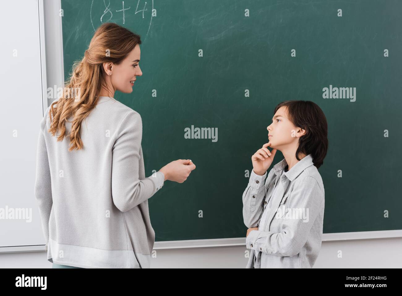 thoughtful schoolboy looking at smiling teacher near chalkboard Stock ...