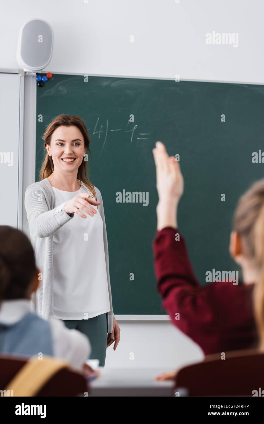 happy teacher pointing at schoolgirl raising hand on blurred foreground ...