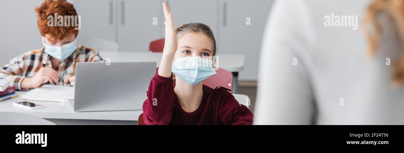 schoolgirl in medical mask raisin hand during lesson near teacher on ...