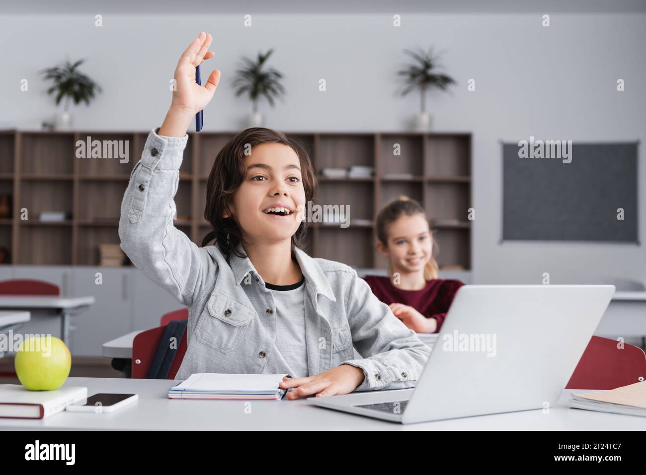 Cheerful boy raising hand near notebook and devices in classroom Stock ...