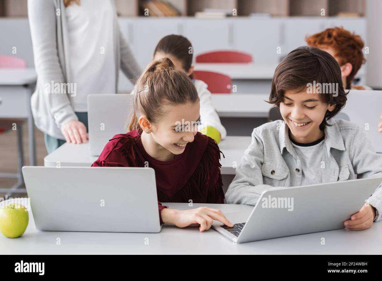 cheerful kids typing on laptop near classmates and teacher on blurred ...
