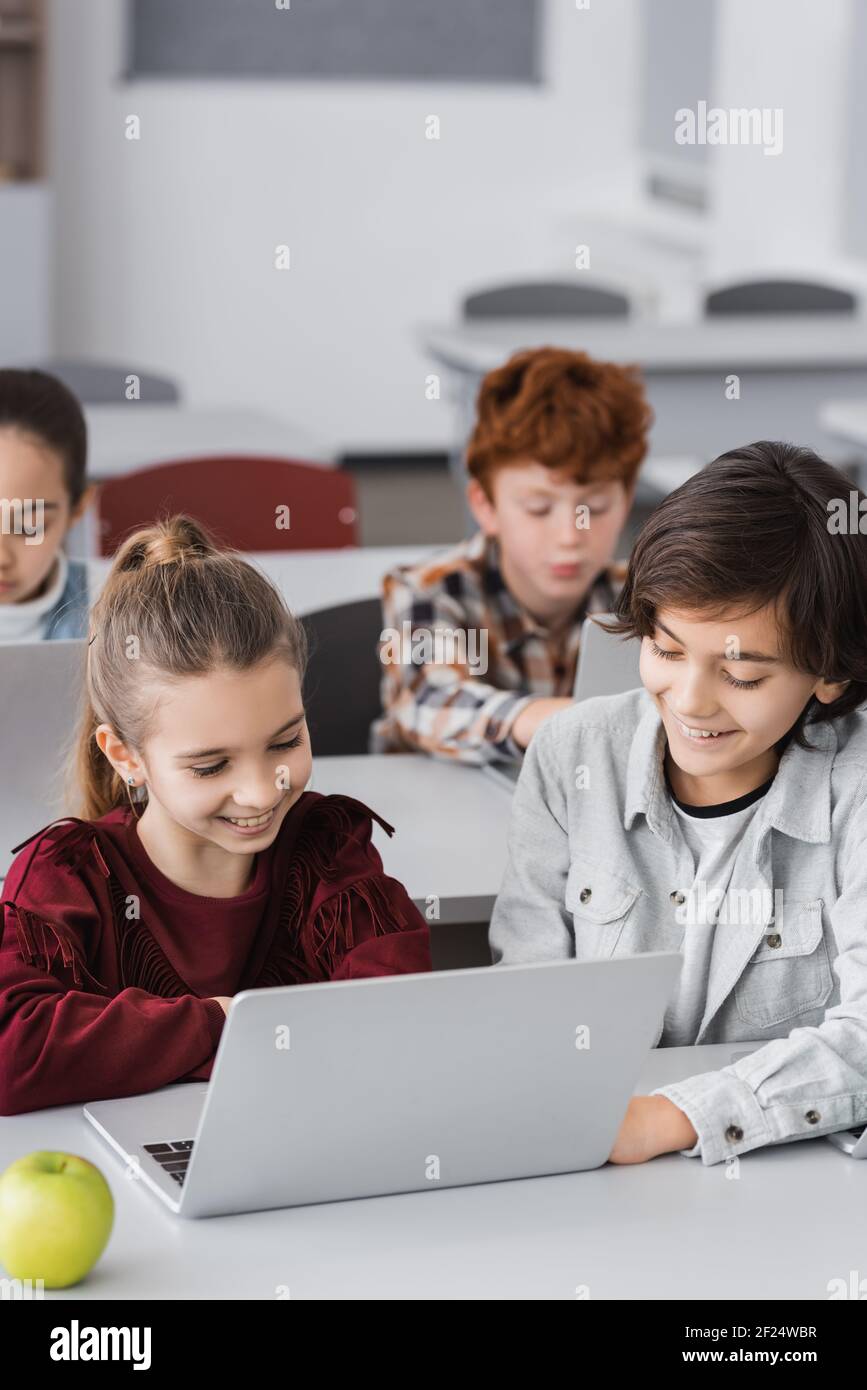 cheerful classmates near laptops, apple and pupils on blurred ...