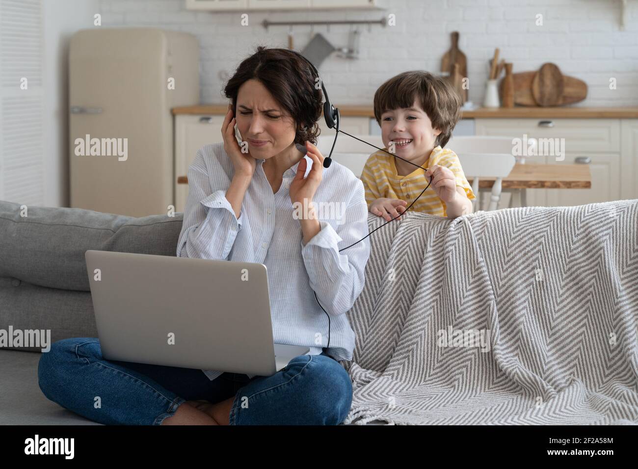 Exhausted mother sitting on couch at home, working on laptop, child ...