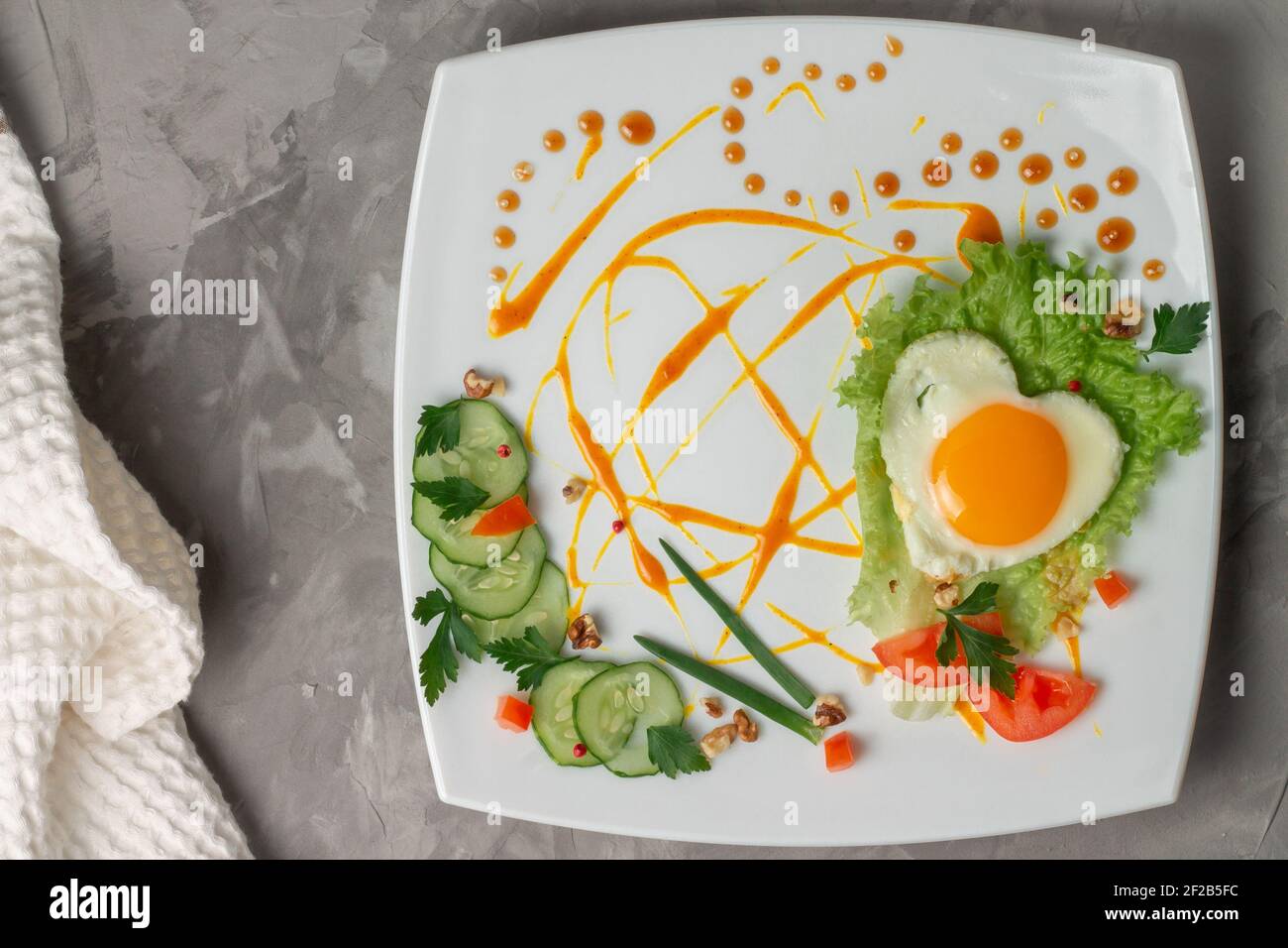 Heart shaped fried egg with vegetables, herbs and sauce on square white ...