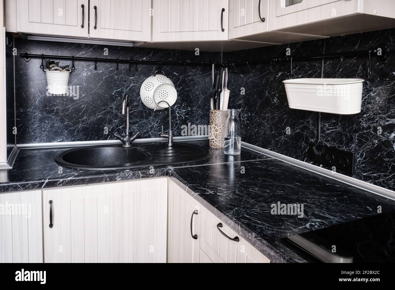 A corner with a sink in a white kitchen with a black marble countertop ...
