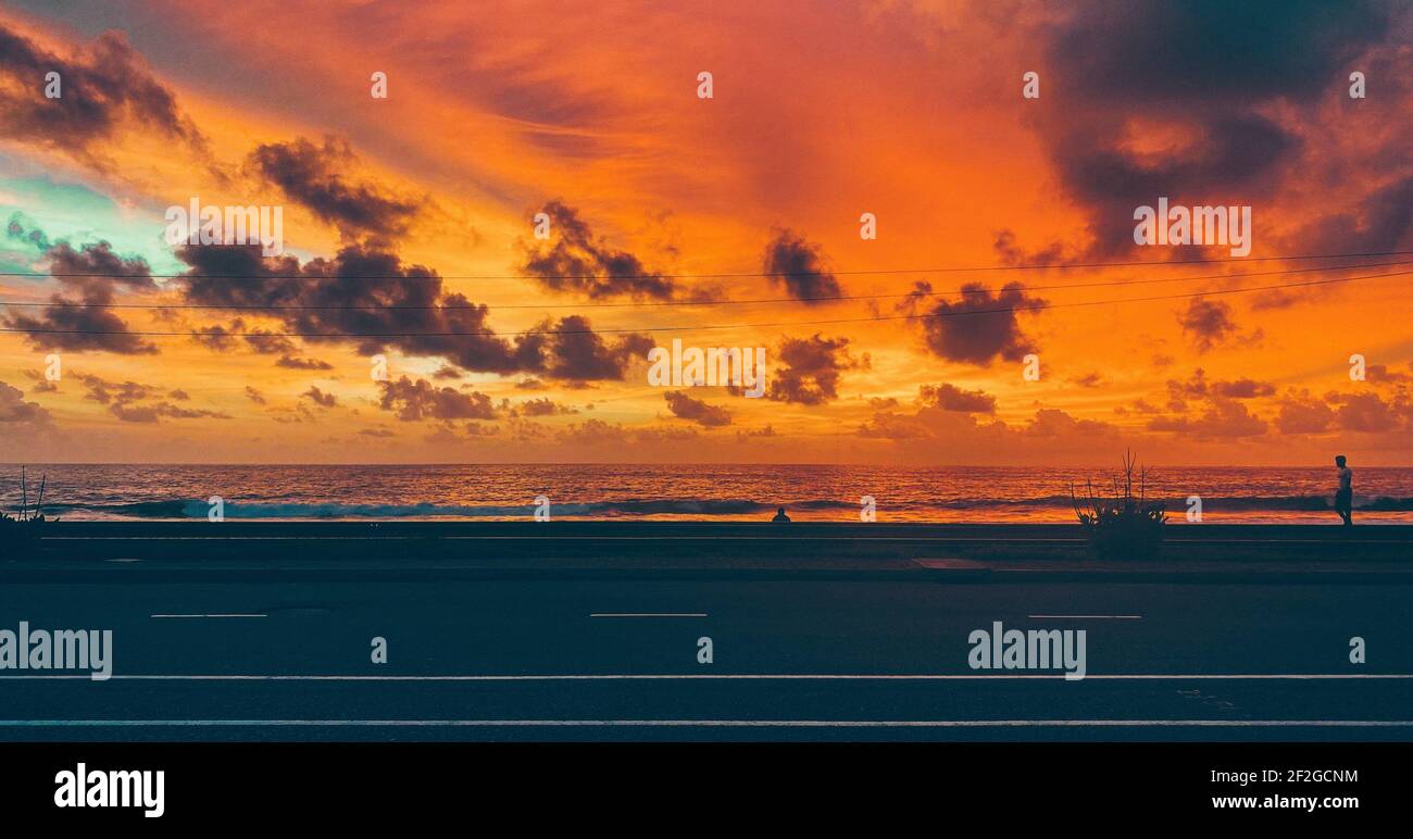 A silhouette of a man walking a paved waterfront highway under a majestic sunset cloudscape ...