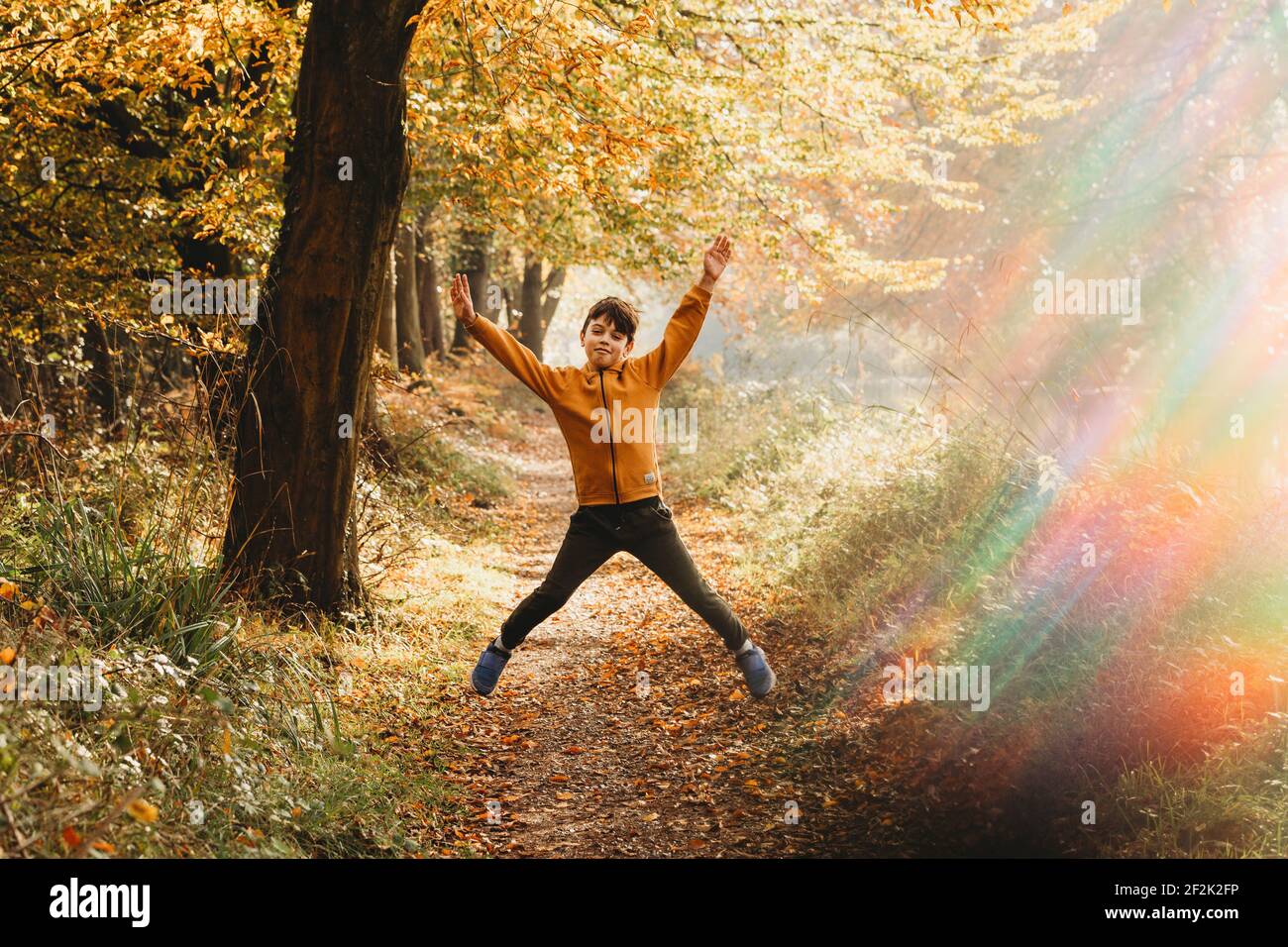 Boy leaping in the air on path under tree with rainbow light flare ...