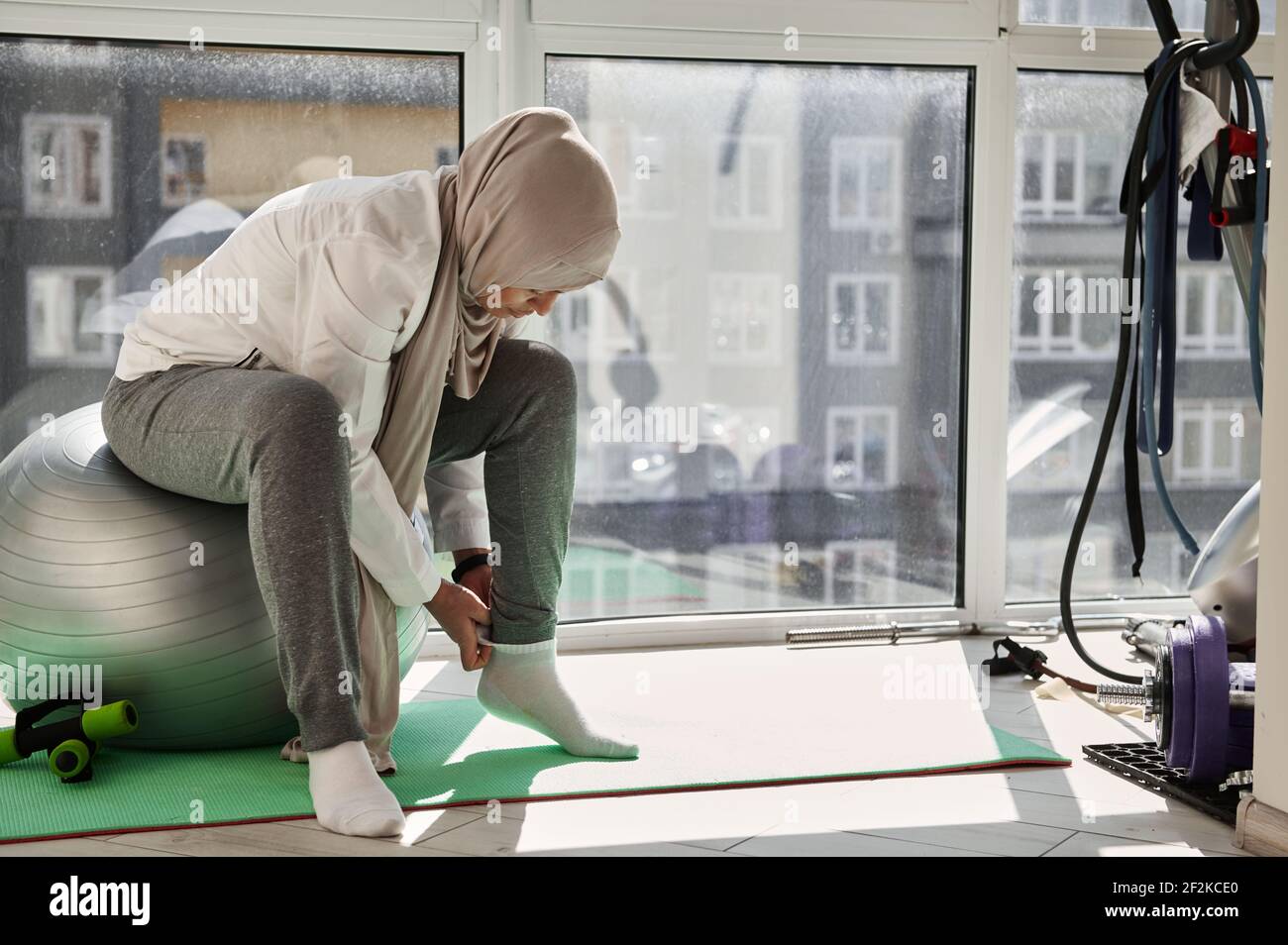 Athletic Arab Muslim woman in hijab sits on a fitness ball and puts on ...